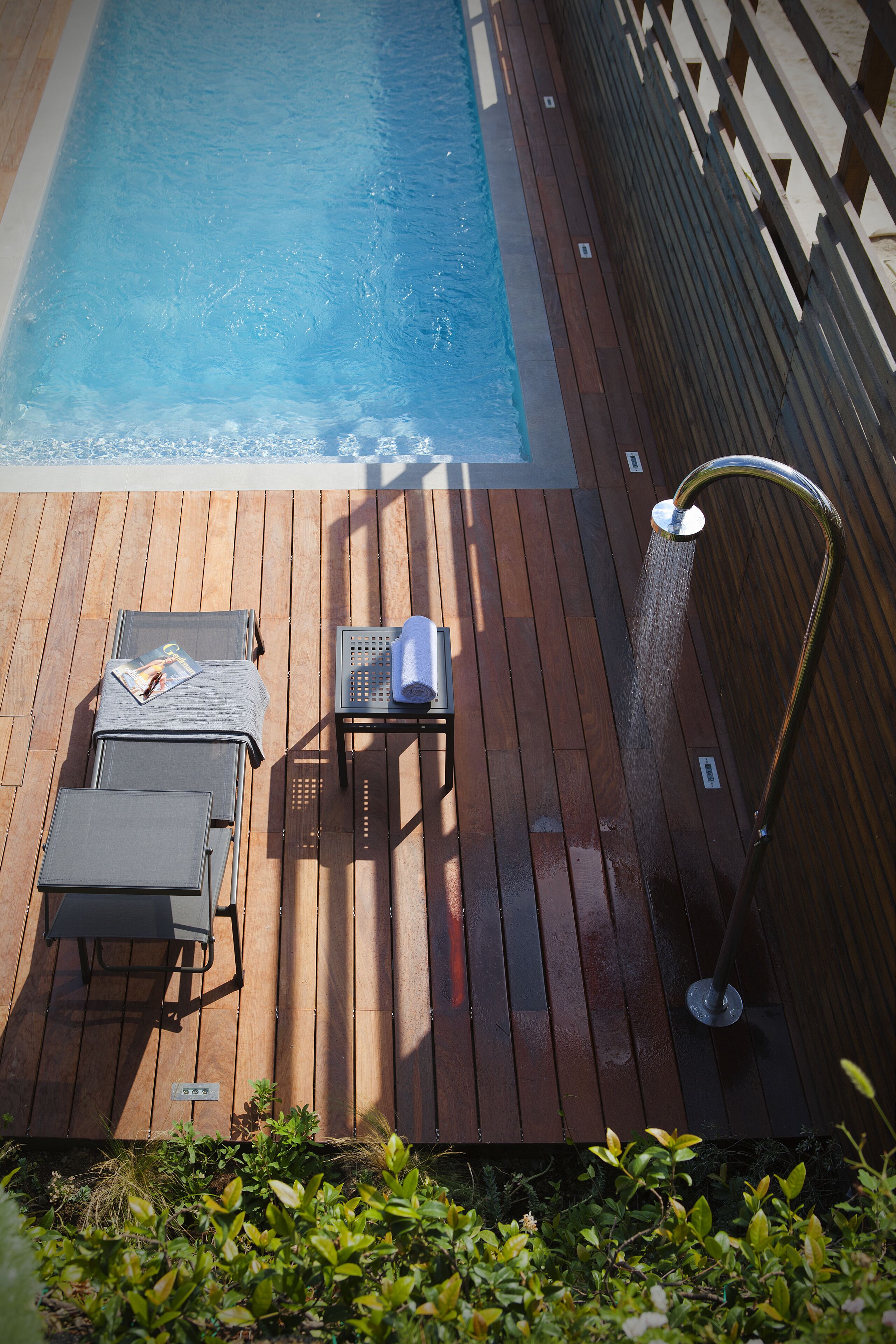 Um deck de madeira com piscina, uma cadeira de descanso, uma mesa com revista e um chuveiro.