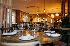 Interior de restaurante com mesas redondas preparadas para refeições, com copos de vinho vazios e pratos, e luminárias pendentes.