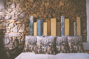 Uma cama com lençol branco, parede de pedra e cabeceira de madeira com tinta descascada.