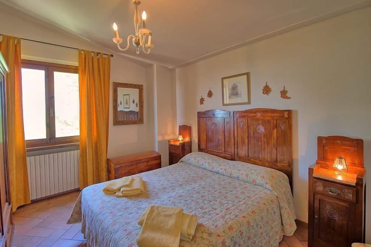 Quarto com cama, cabeceira de madeira, mesas de cabeceira, um candelabro e janela com cortinas.