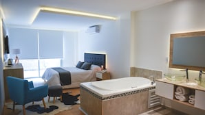 Quarto de hotel moderno com uma cama grande, área de estar separada com uma poltrona, banheira, penteadeira com espelho e uma janela ampla que proporciona iluminação natural.