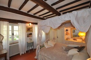 Quarto com cama de dossel, mesa de cabeceira, cômoda, espelho e janela com cortinas.