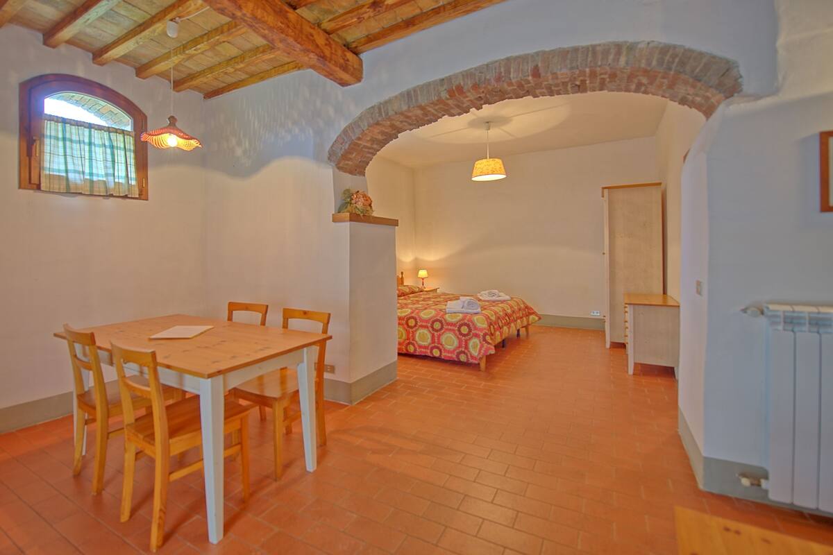 Um quarto com uma mesa de jantar de madeira e cadeiras, uma cama com colcha estampada, um lustre e um arco de tijolos.