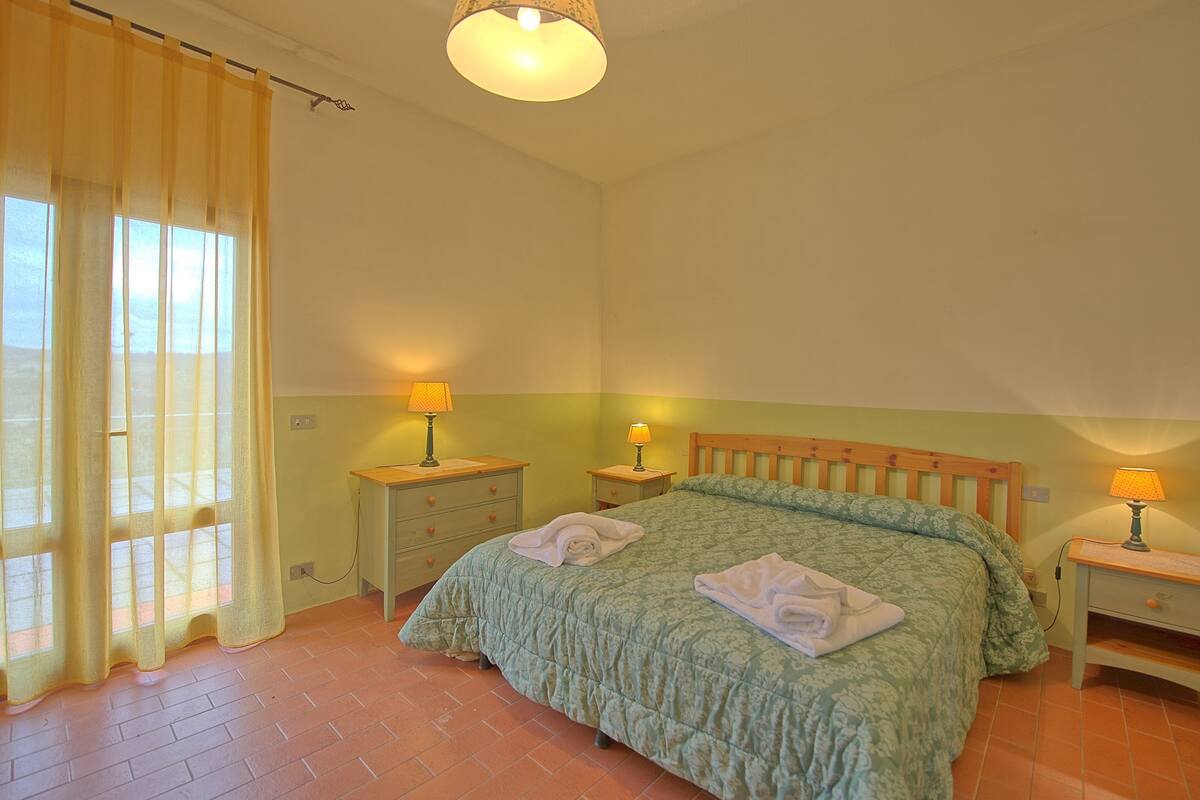Quarto com colcha verde, duas mesas de cabeceira com luminárias, cabeceira de madeira e janela com cortinas amarelas.