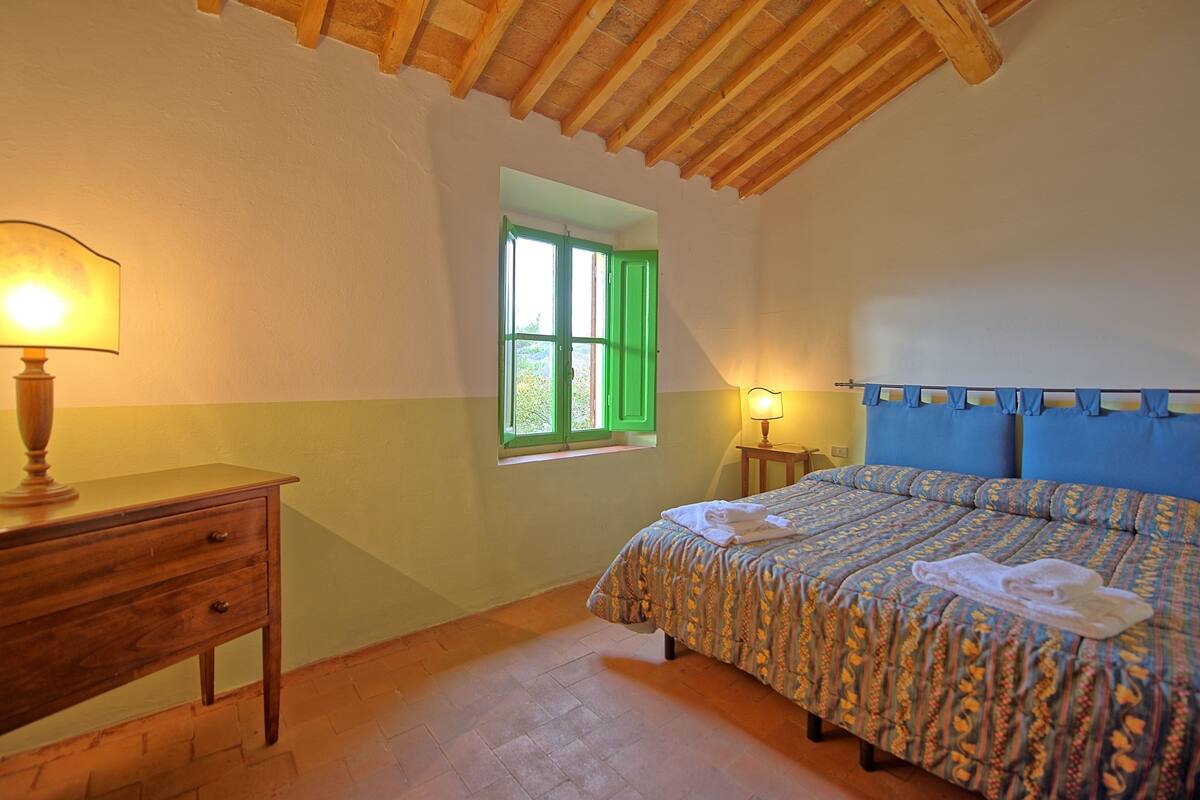 Quarto com teto de madeira, cama com colcha estampada, mesa de cabeceira com abajur e janela com persianas verdes.