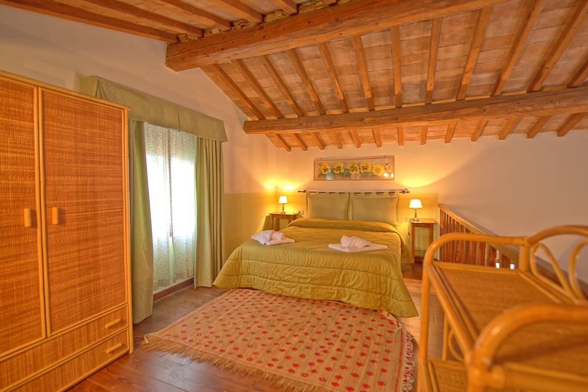 Quarto com teto de madeira, cama com roupa de cama verde, duas luminárias de cabeceira, guarda-roupa de madeira e tapete floral.