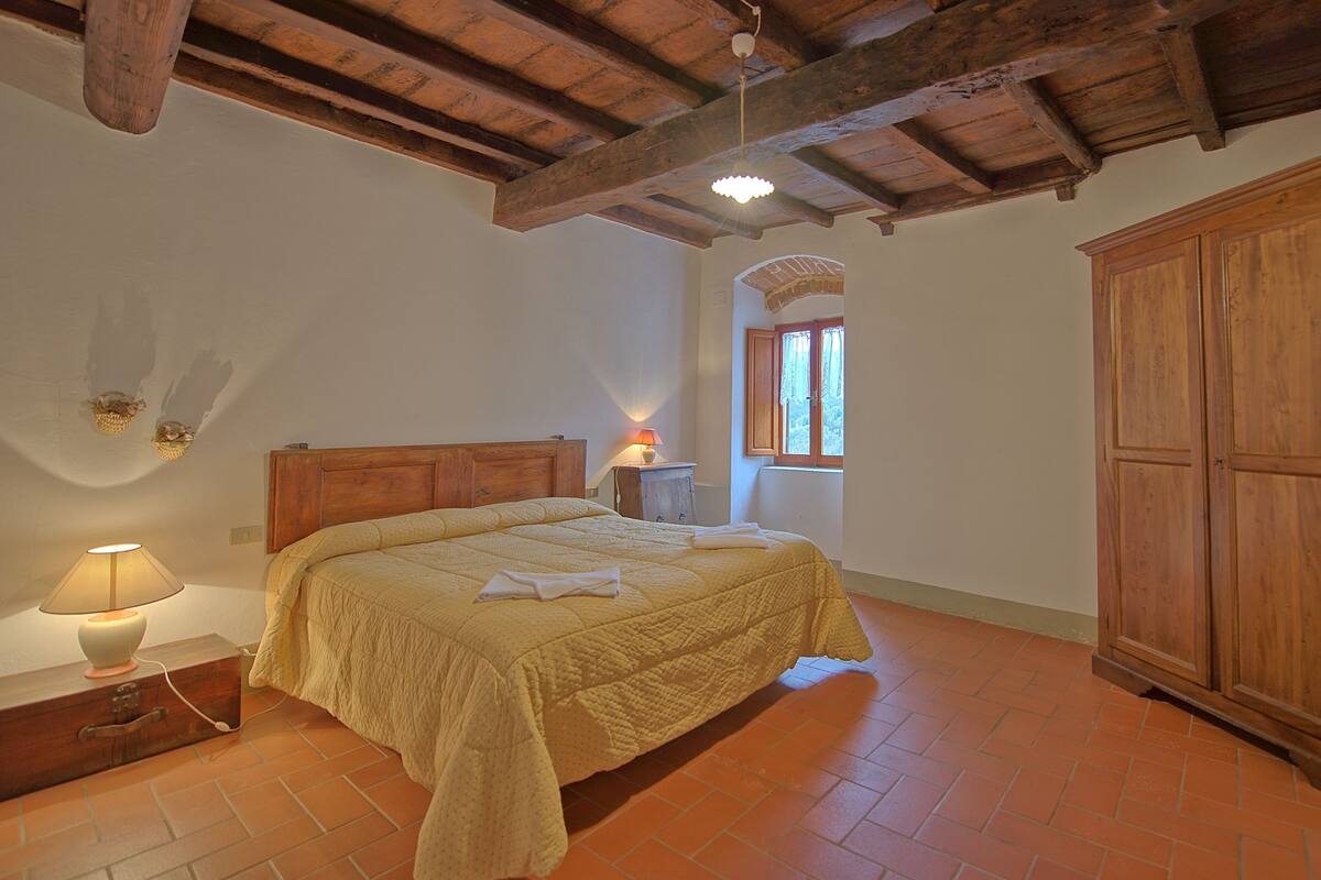 Quarto com cabeceira de madeira, cama com colcha bege, mesa de cabeceira com abajur e guarda-roupa de madeira.