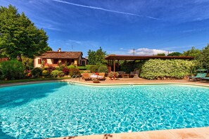 Piscina com espreguiçadeiras e um gazebo, rodeada por vegetação exuberante e uma casa.
