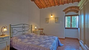 Quarto com cama, duas mesas de cabeceira, um criado-mudo, teto de madeira e janela com vista para o verde.
