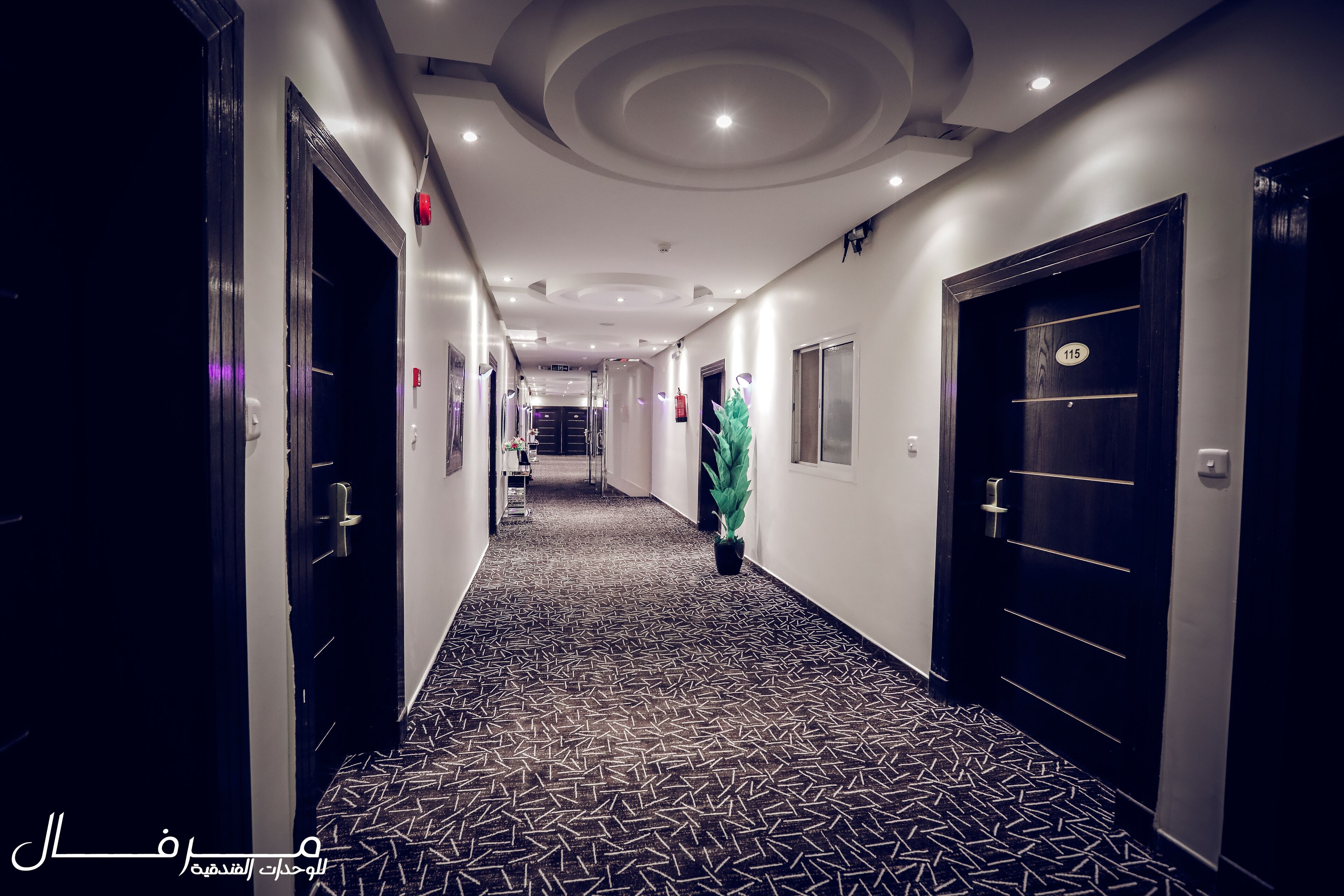 Corredor de hotel com carpete estampado, portas escuras e iluminação embutida.