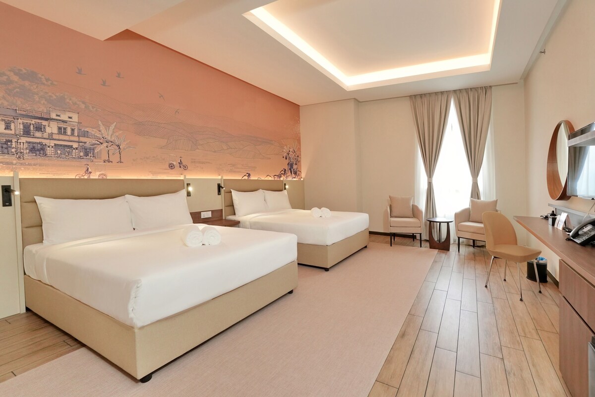 Quarto de hotel com duas camas, uma escrivaninha, uma cadeira, uma mesa pequena e um espelho grande.