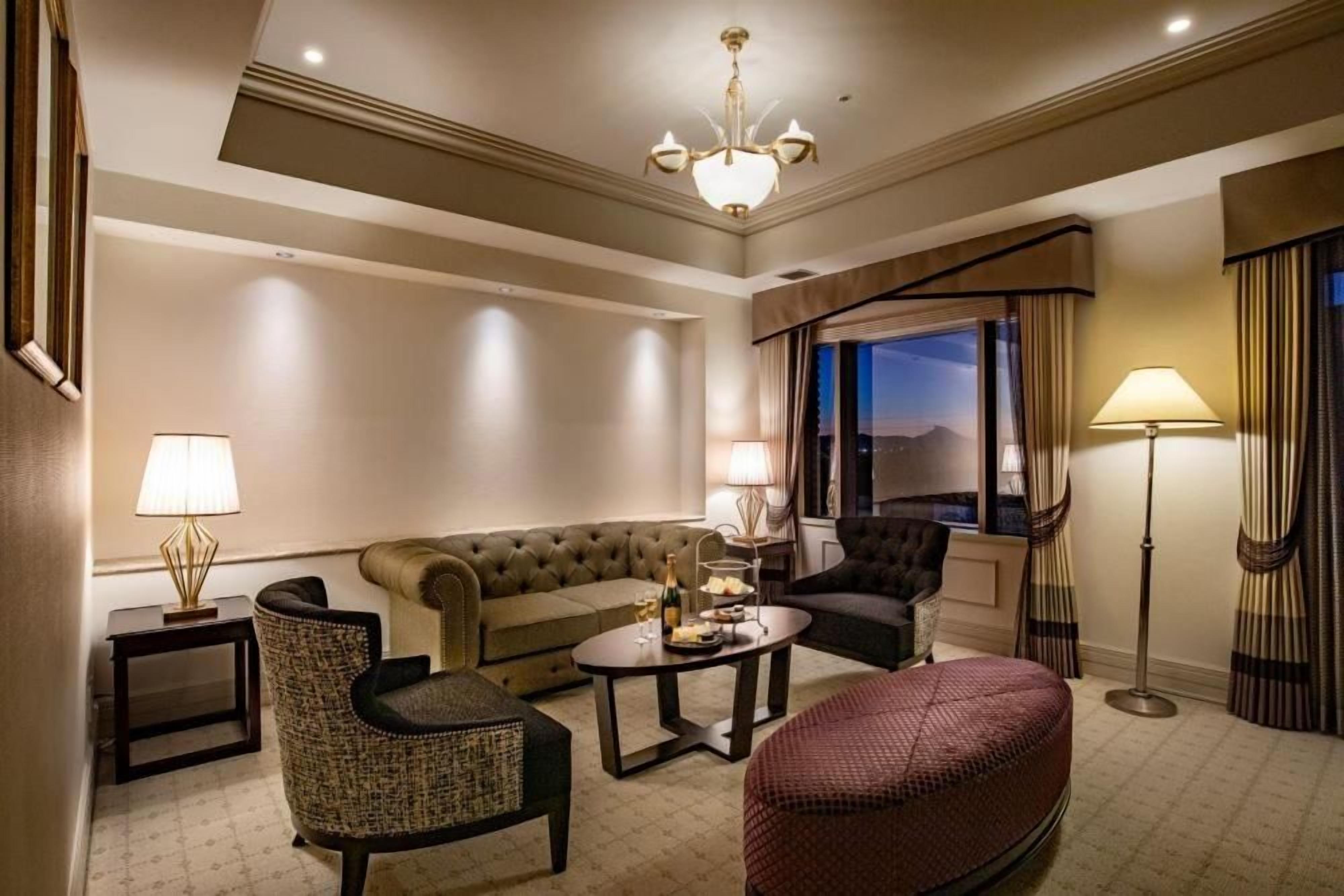 Quarto de hotel com sofá, poltronas, mesa de centro, luminária de chão e vista para o exterior.