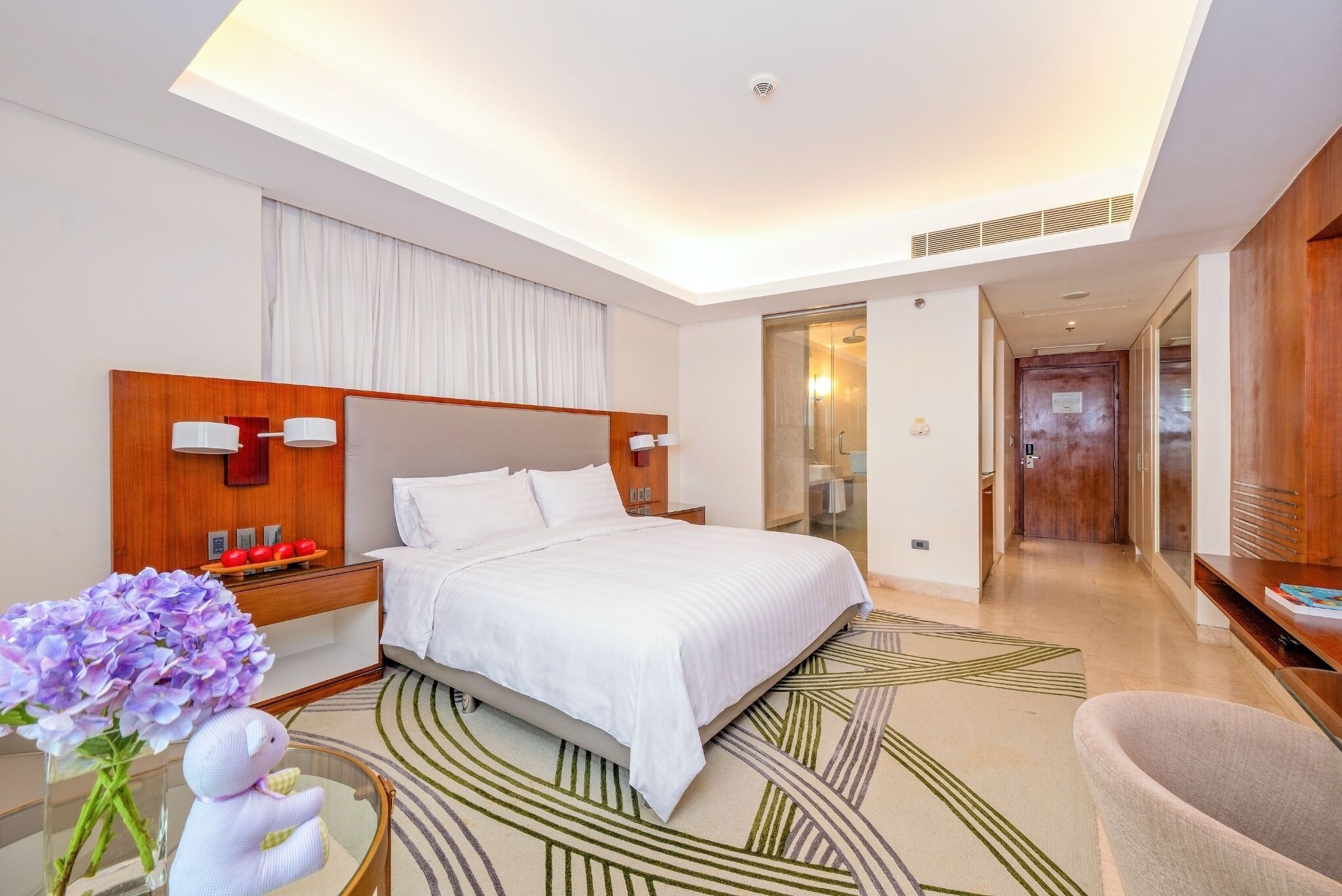 Quarto de hotel moderno com uma cama grande, mesas de cabeceira, uma mesa de centro de vidro com um vaso de flores e vista para o corredor.