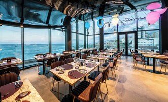 Um restaurante com mesas e cadeiras dispostas para refeições, amplas janelas com vista para o mar e edifícios, e luminárias pendentes.