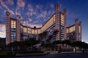 Um hotel com vários andares, fachada curva, múltiplas varandas e um pátio ajardinado com palmeiras.