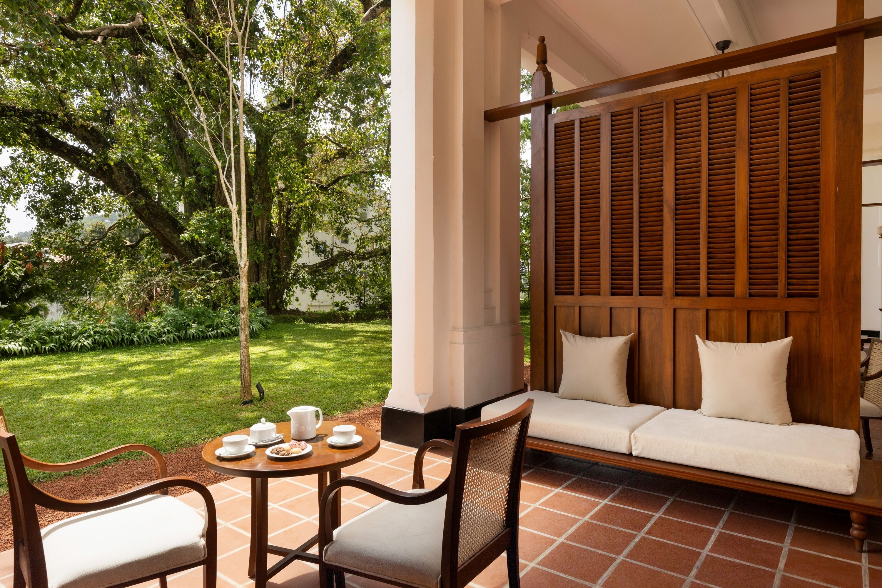 Uma área de pátio com um banco de madeira, uma mesa com chaleira e xícaras, e vista para um jardim exuberante.