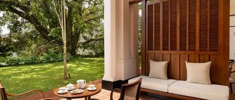 Uma área de pátio com um banco de madeira, uma mesa com chaleira e xícaras, e vista para um jardim exuberante.