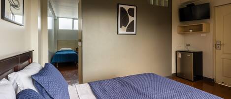 Quarto de hotel com cama, televisão, pequeno frigorífico e quadro na parede.