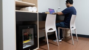 Uma pessoa sentada em uma mesa com um laptop, uma geladeira com bebidas e uma estante com toalhas dobradas.