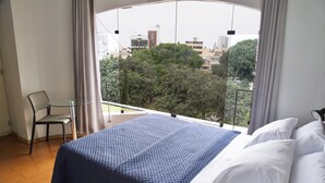 Quarto com cama, cadeira e vista para a cidade através de grandes janelas.