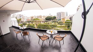 Uma varanda com vista para a paisagem urbana, com duas cadeiras de vime e uma pequena mesa redonda com um copo de suco de laranja.