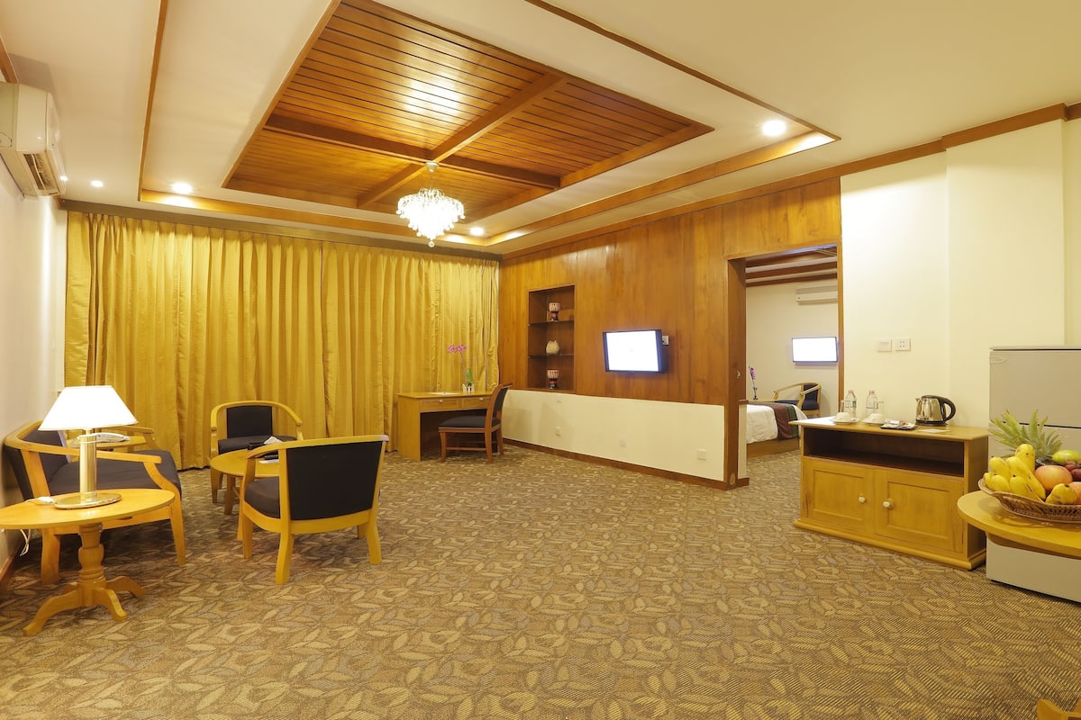 Quarto de hotel com teto de madeira, lustre, secretária com televisão de tela plana, pequena copa e área de jantar com cesta de frutas.