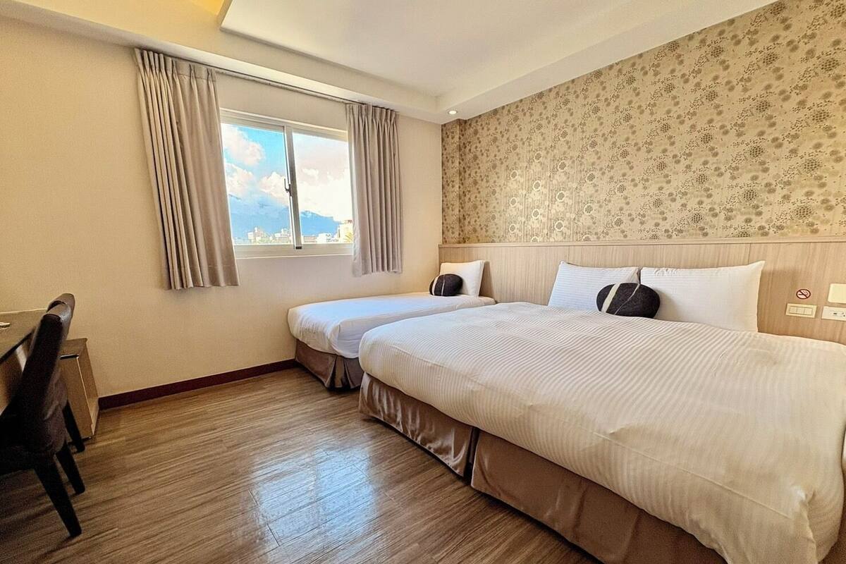 Quarto de hotel com duas camas, piso de madeira, janela com cortinas e papel de parede estampado.