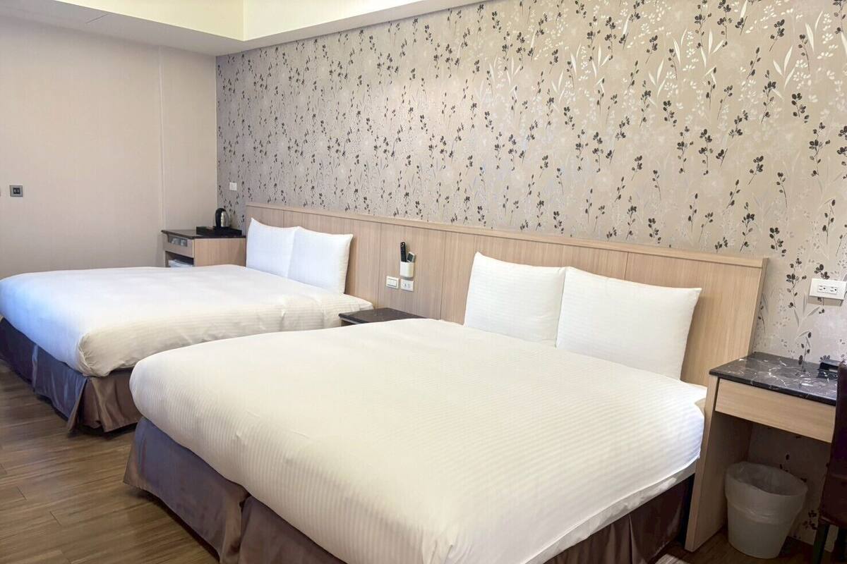 Quarto de hotel com duas camas, cabeceiras de madeira, pequenas mesas de cabeceira com luminárias e uma parede com padrão floral.