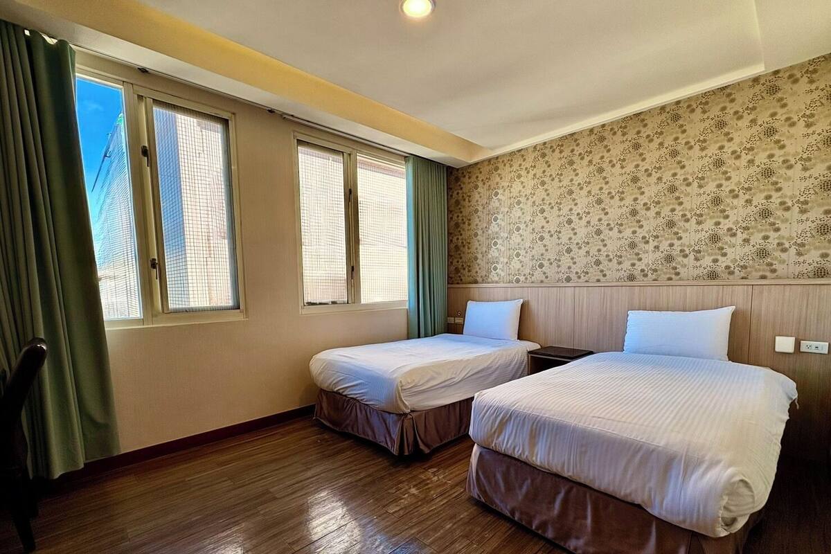 Quarto de hotel com duas camas, piso de madeira, papel de parede estampado e vista para edifícios pelas janelas.
