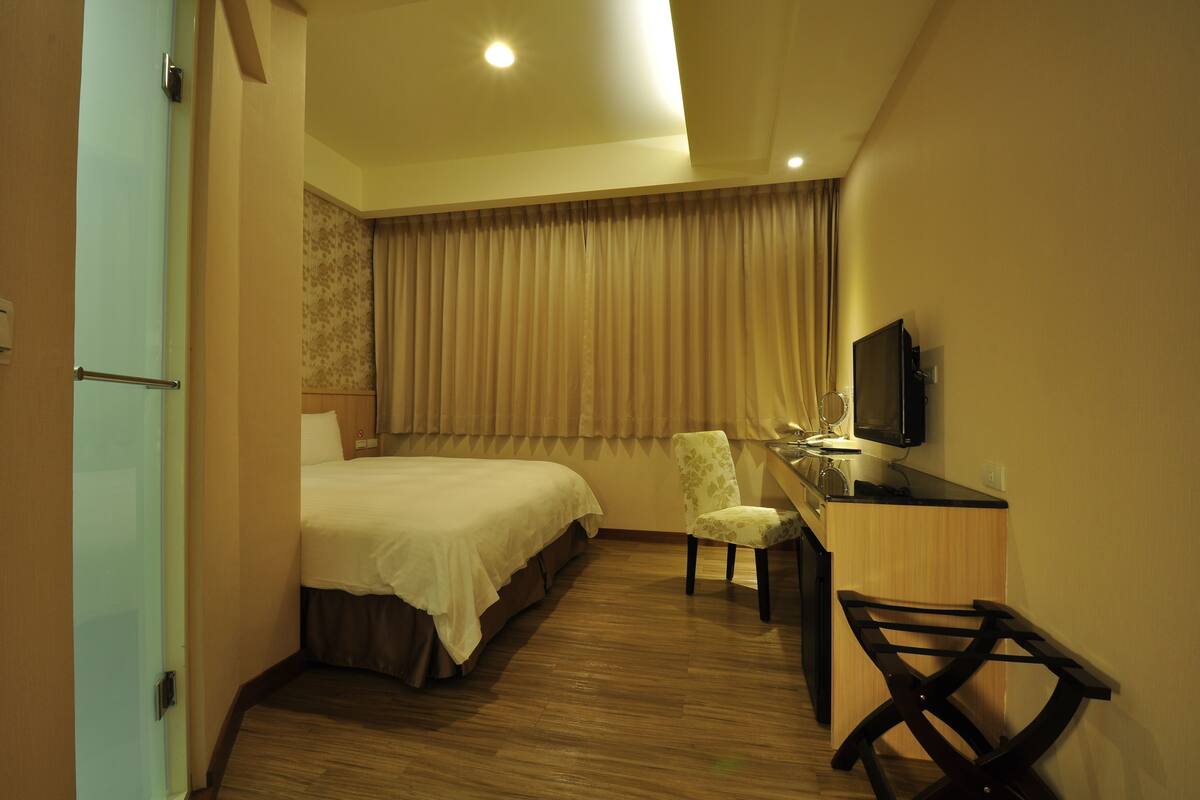 Quarto de hotel com cama, escrivaninha, cadeira, televisão e cortinas.