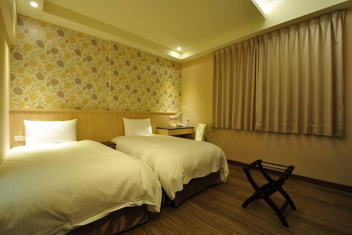 Quarto de hotel com duas camas, uma cabeceira de madeira, uma pequena mesa com abajur e uma janela com cortinas.