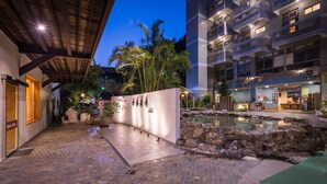 Pátio de hotel com caminho de pedra, um pequeno espelho d'água e uma área coberta com teto de madeira.