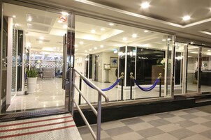 Entrada de hotel com porta de vidro, barreira de corda azul e escadaria de metal que dá acesso ao interior.