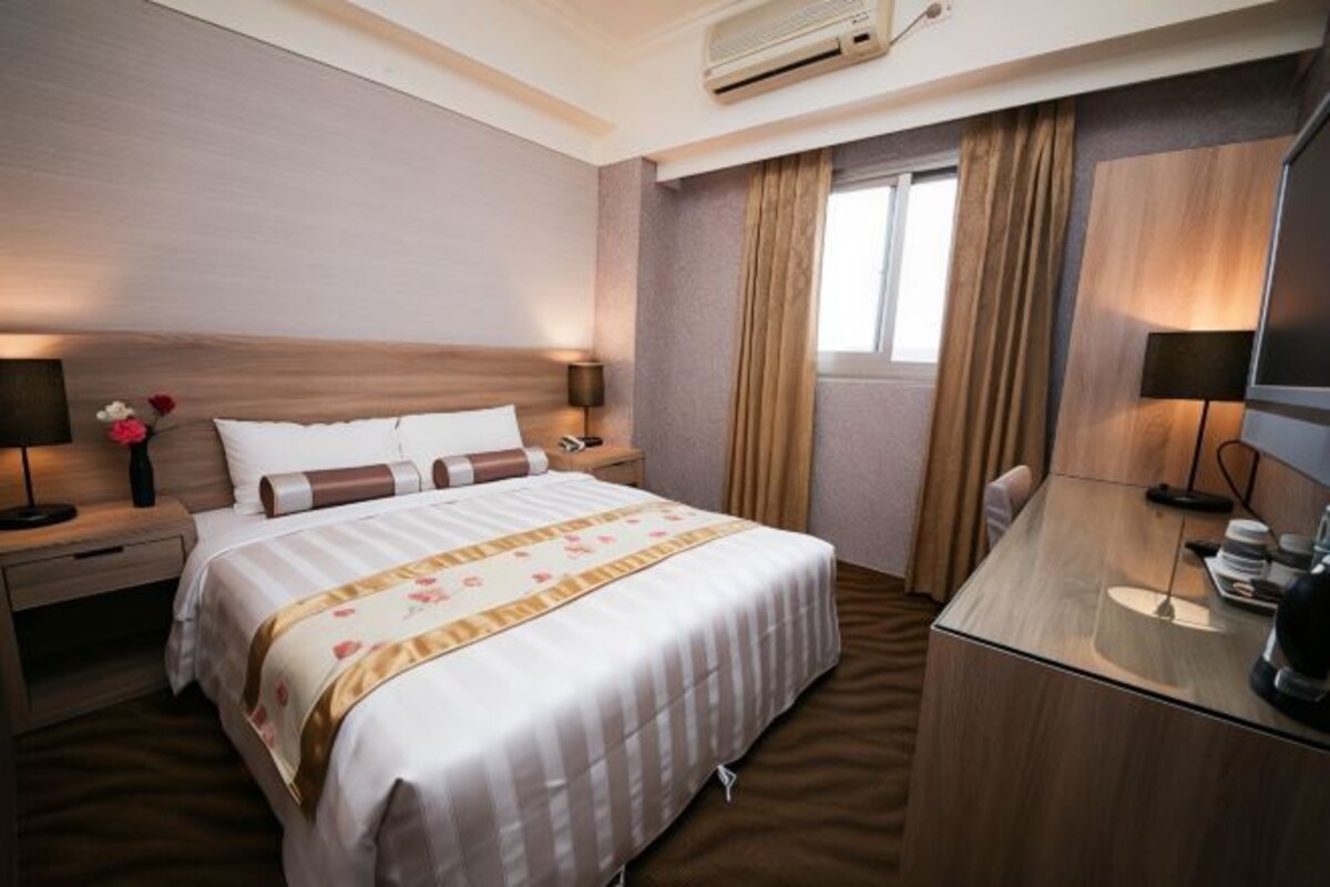 Quarto de hotel com cama, mesas de cabeceira, escrivaninha e janela com cortinas.