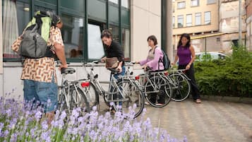 Um grupo de pessoas perto de bicicletas estacionadas em frente a um edifício, com um caminho de tijolos e flores roxas ao primeiro plano.