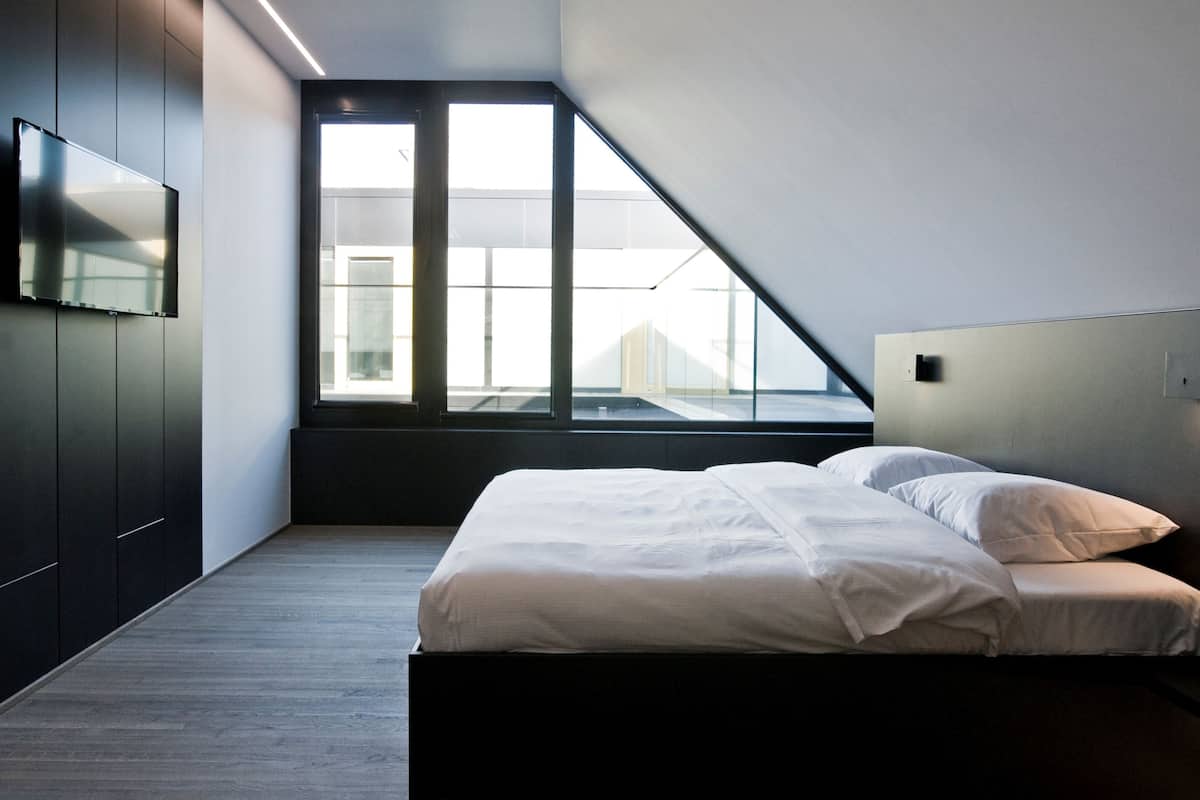 Quarto moderno com teto inclinado, uma janela grande e cama com roupa de cama branca.