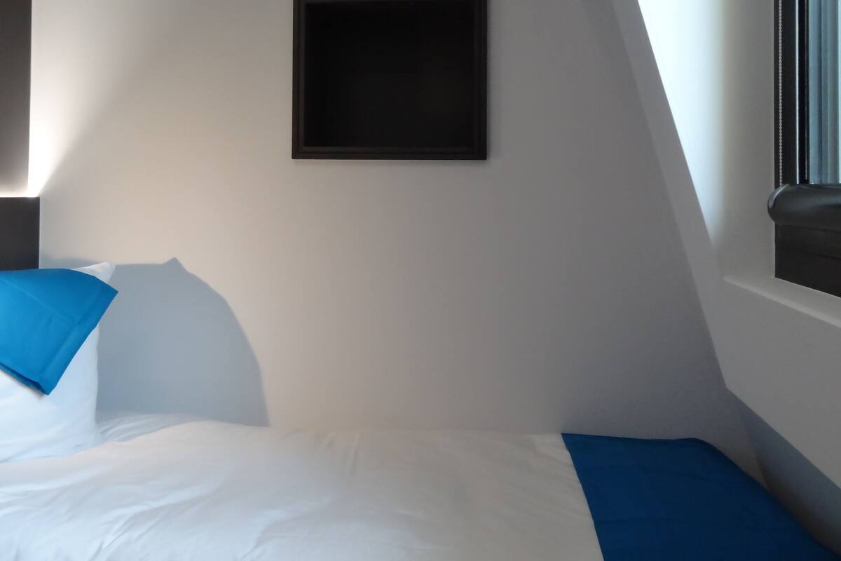 Quarto de hotel moderno com cama, uma janela pequena e luminária fixada na parede.
