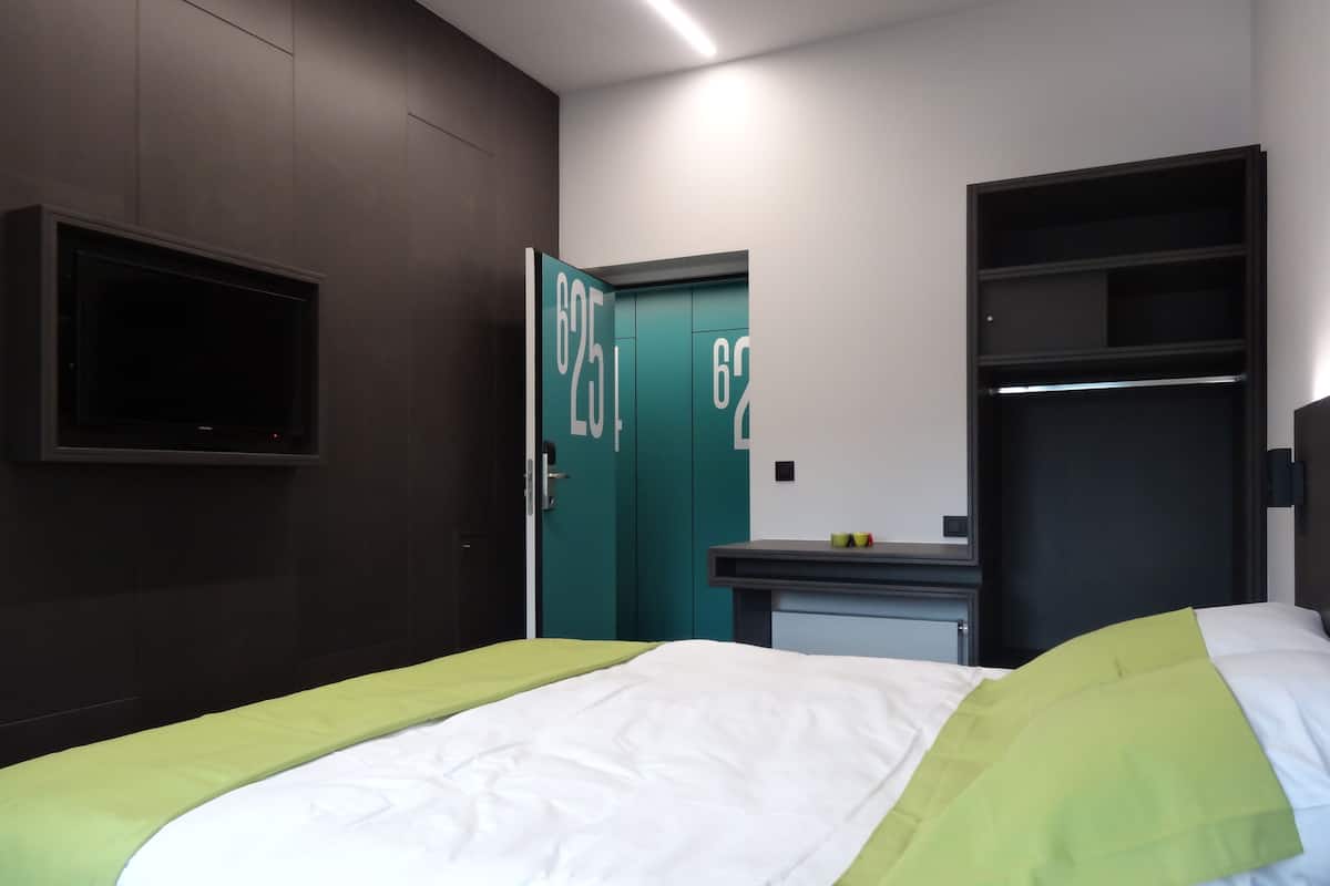 Quarto de hotel com cama, televisão na parede, guarda-roupa e pequena mesa de trabalho com espelho.