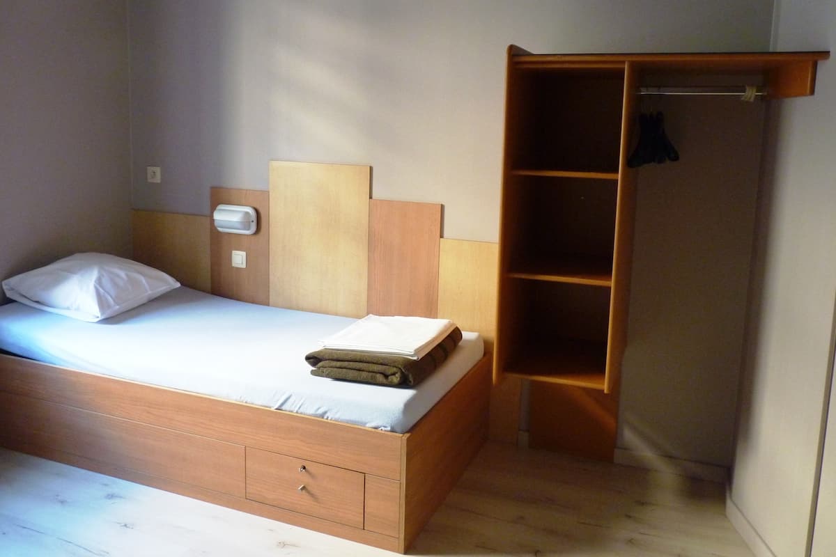 Uma cama de solteiro com cabeceira de madeira, mesa de cabeceira e guarda-roupa embutido com prateleiras.