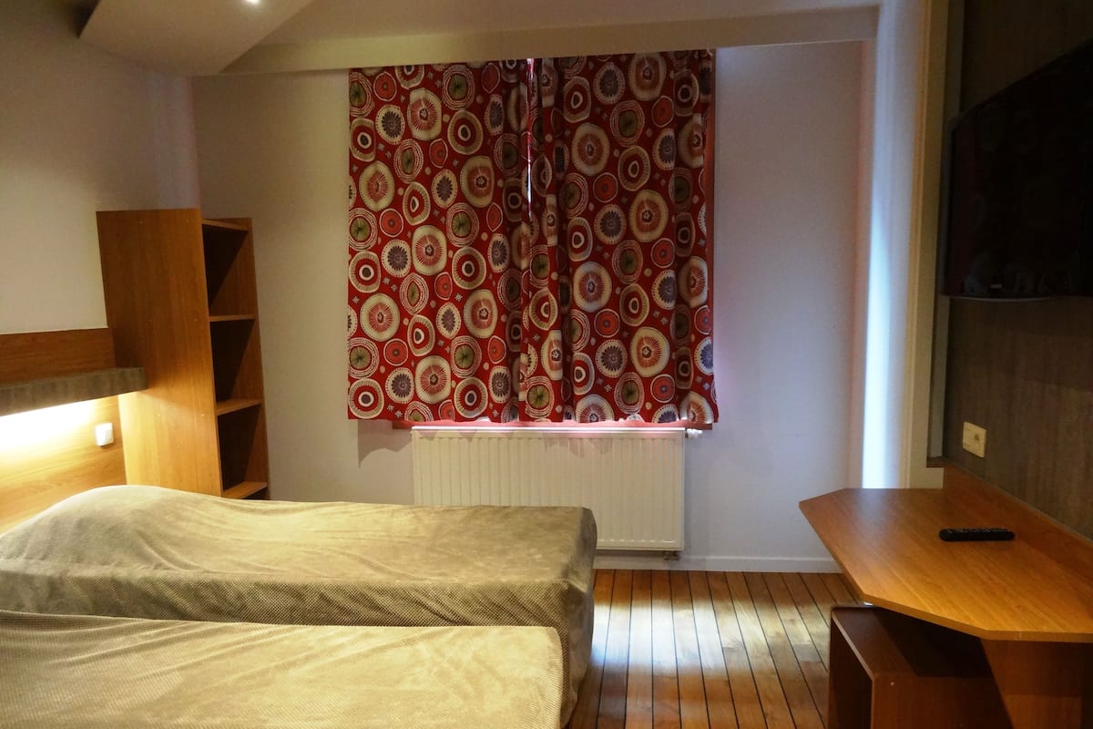 Quarto com cama, mesa de madeira, radiador e janela com cortinas estampadas.