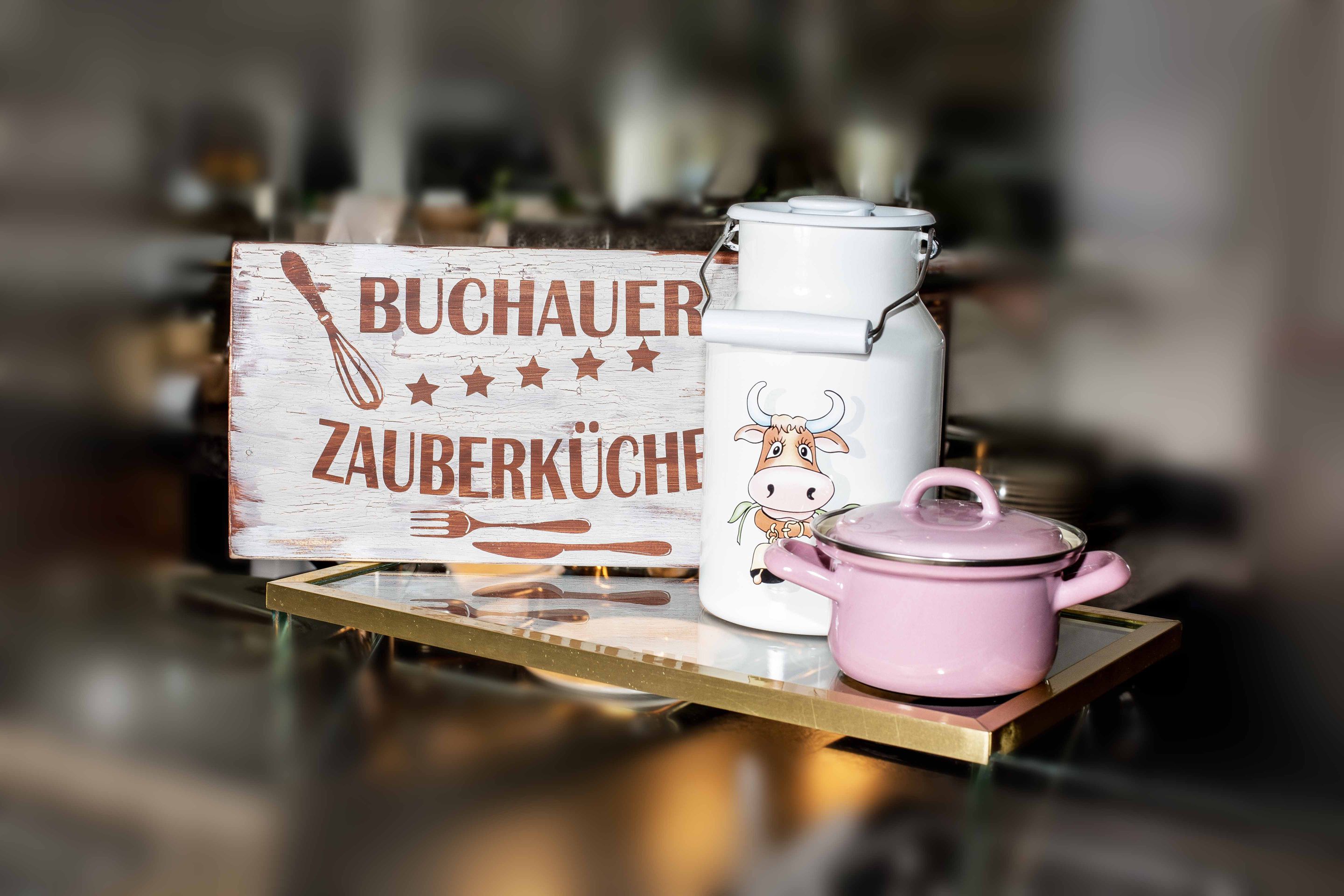 Uma bancada de cozinha com uma placa que diz "BUCHAUER ZAUBERKÜCHE" e uma panela rosa com tampa.