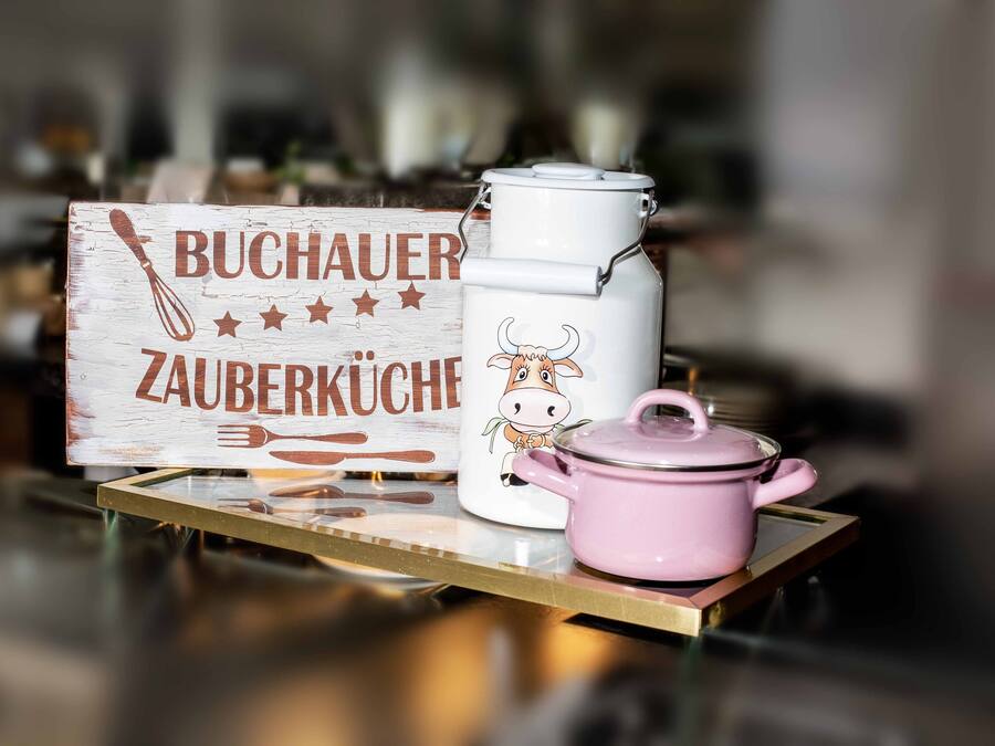 Uma bancada de cozinha com uma placa que diz "BUCHAUER ZAUBERKÜCHE" e uma panela rosa com tampa.