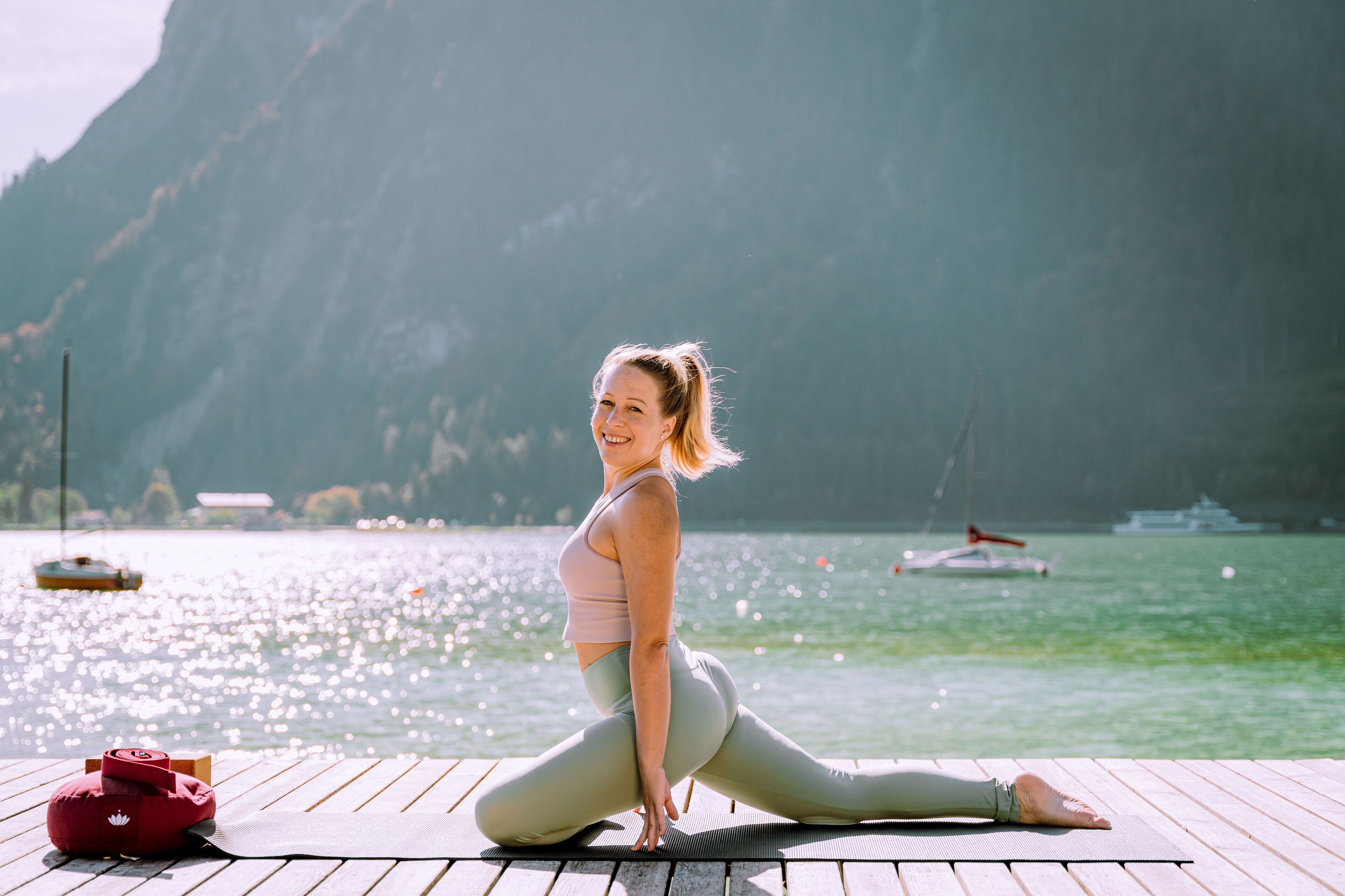 Uma pessoa fazendo uma postura de yoga em um deck de madeira perto de um corpo d'água, com barcos ao fundo.