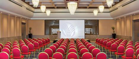 Sala de conferências com fileiras de cadeiras vermelhas, uma tela grande e lustres.