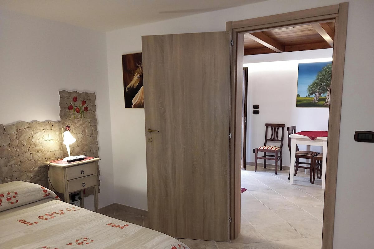 Quarto com porta de madeira, luminária de cabeceira, cama com roupa de cama floral, parede de pedra e um quadro de um cavalo.