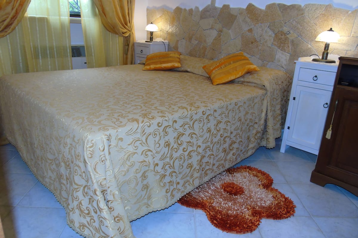 Quarto com cama, duas mesas de cabeceira, um criado-mudo, abajur e parede de pedra.