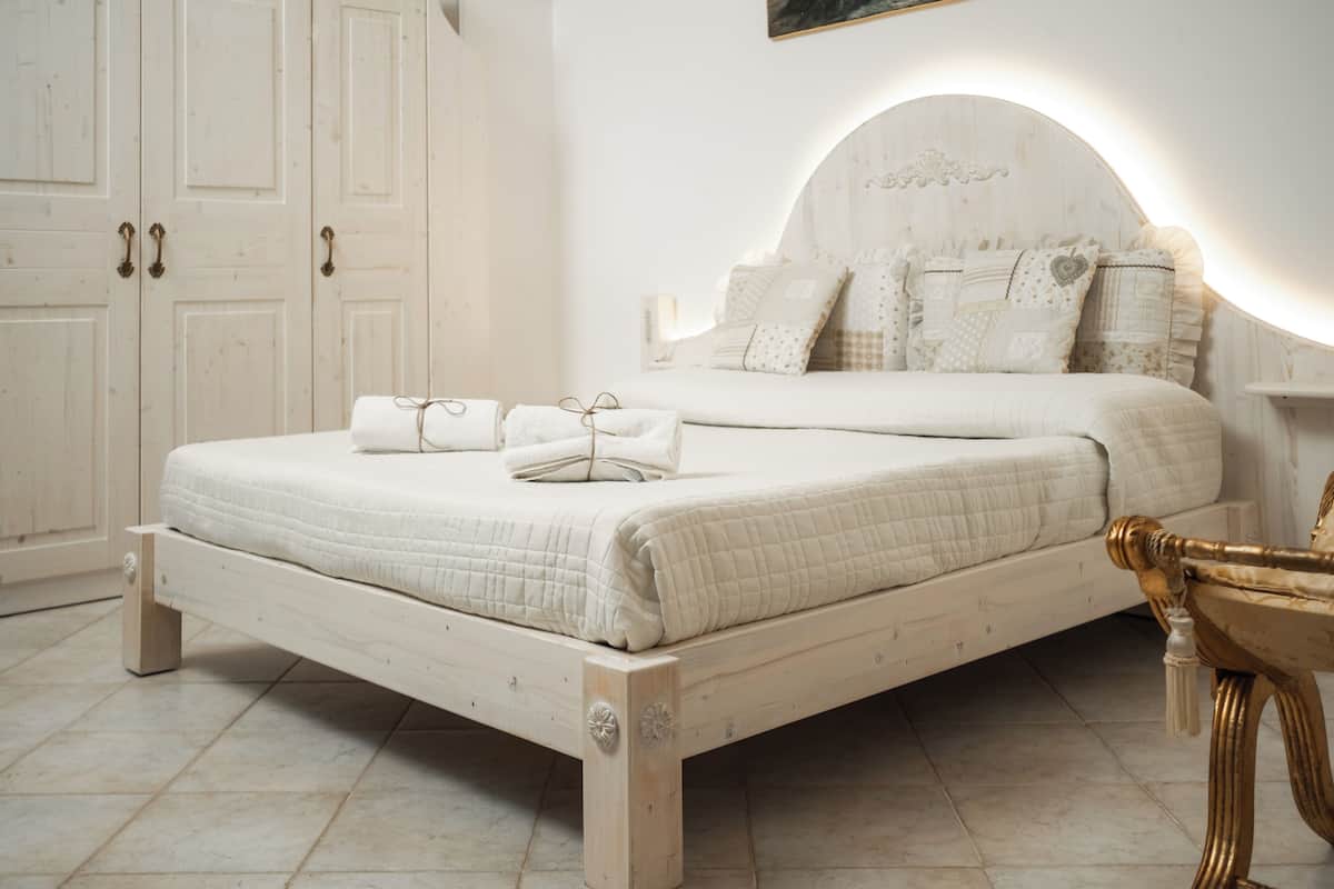 Quarto com cama de madeira, roupa de cama branca e cabeceira com luzes fixadas na parede.