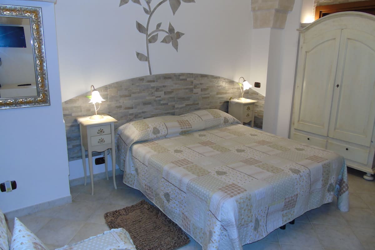 Quarto com cama, mesas de cabeceira, cômoda, espelho e parede com desenho floral.