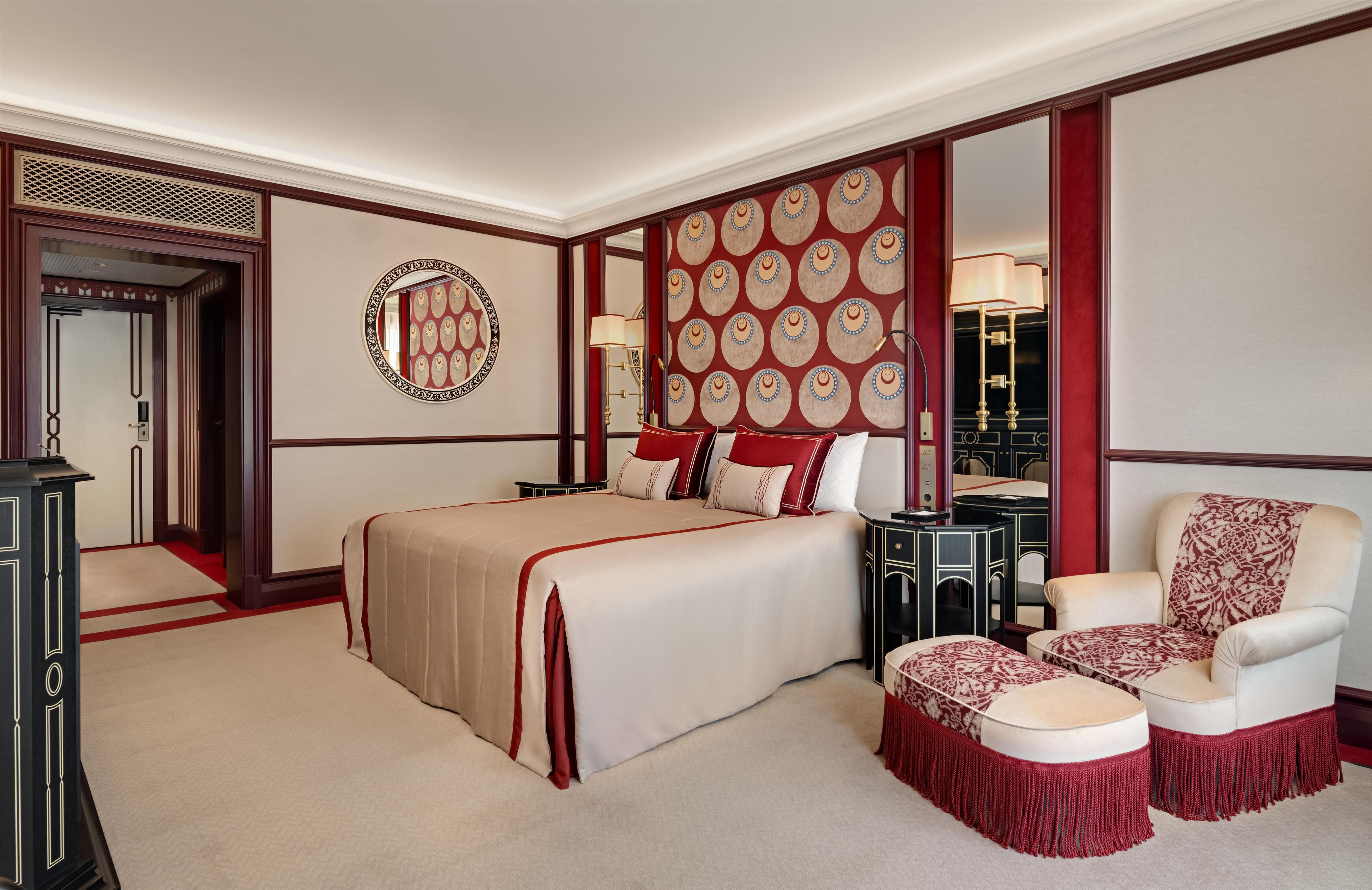 Quarto de hotel com uma cama grande, área de estar com uma poltrona e apoio para os pés, um espelho e uma parede com padrão decorativo.