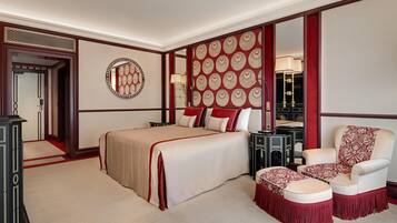 Quarto de hotel com uma cama grande, área de estar com uma poltrona e apoio para os pés, um espelho e uma parede com padrão decorativo.